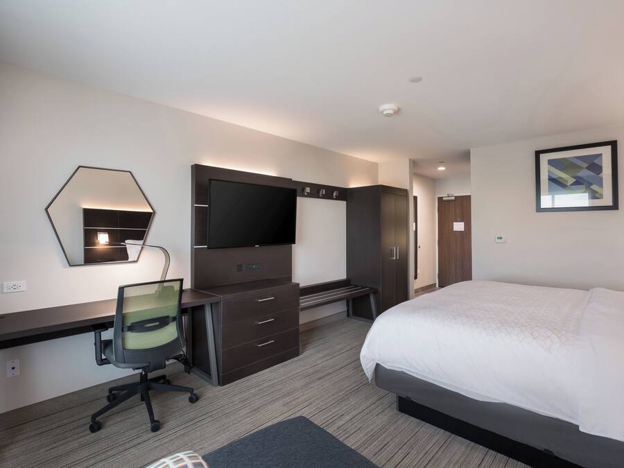 Quarto de hotel com cama, escrivaninha, cadeira, televisão, guarda-roupa e um quadro na parede.