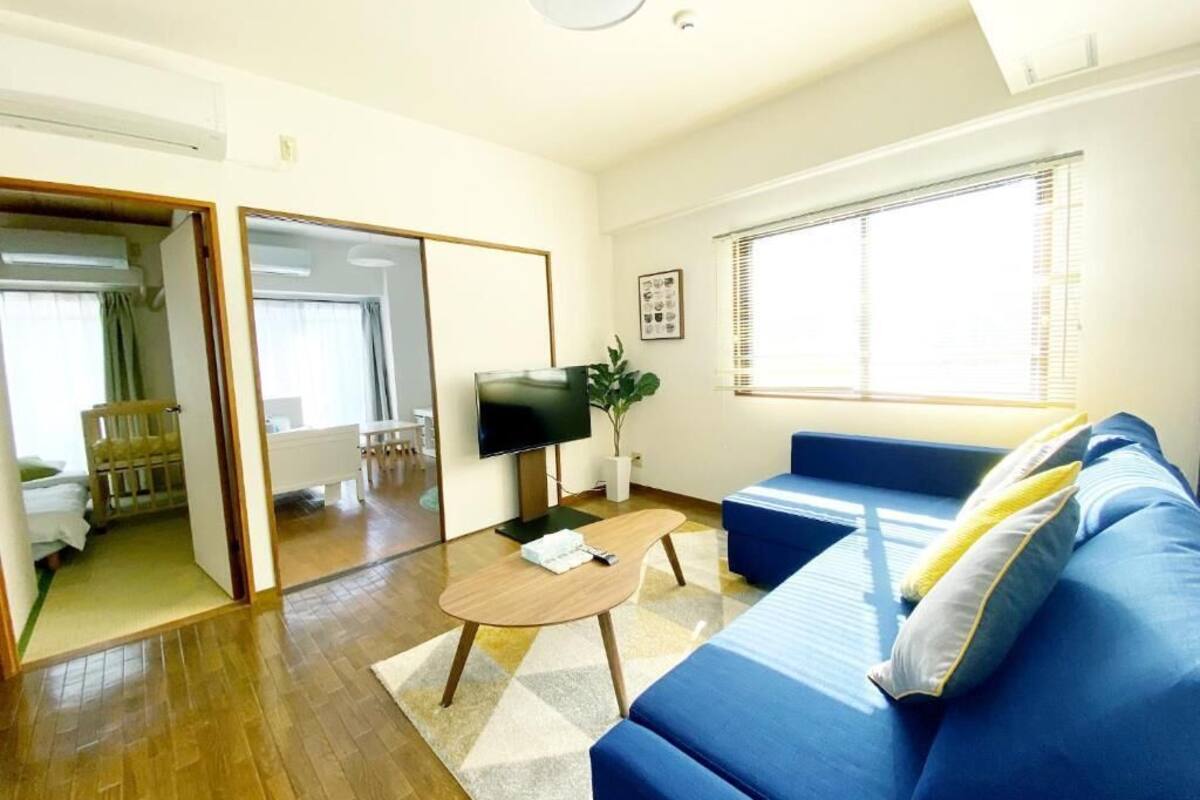 Uma sala de estar com um sofá azul, uma televisão, uma mesa de centro e uma janela grande com persianas.