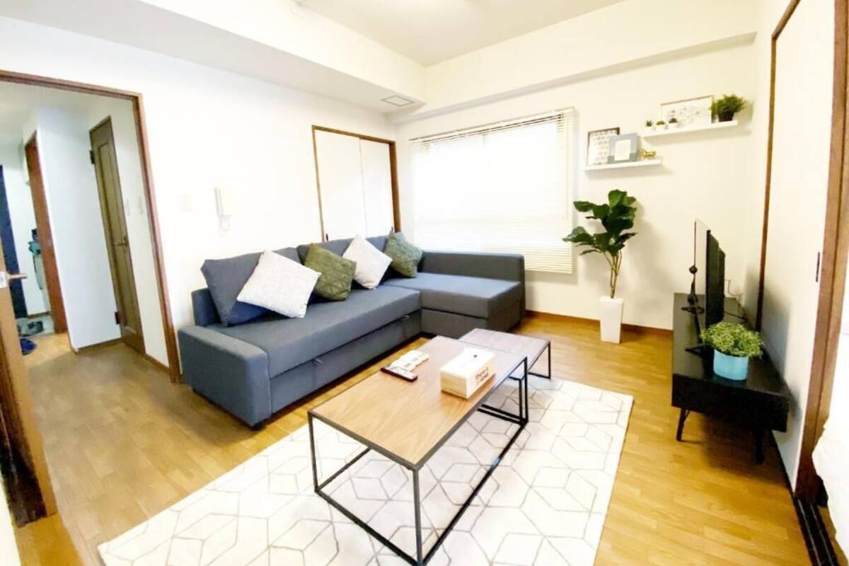 Uma sala de estar moderna com um sofá cinzento, uma mesa de centro de madeira e um tapete branco. Há uma TV sobre um suporte preto e uma planta numa estante.