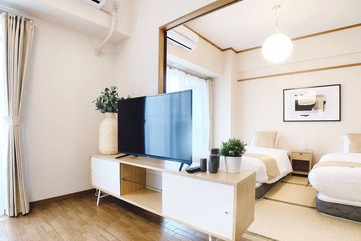 Quarto de hotel moderno com televisão de tela plana em console branco, uma planta em vaso, mesa de cabeceira com abajur, quadro emoldurado na parede e uma janela grande com cortinas.