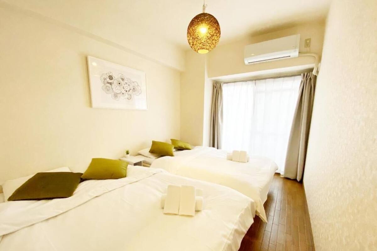 Quarto de hotel com duas camas, luminária pendente, ar condicionado e janela com cortinas.