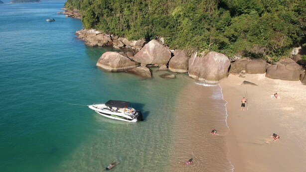 Um barco ancorado perto de uma praia com rochas grandes e uma área arborizada ao fundo.