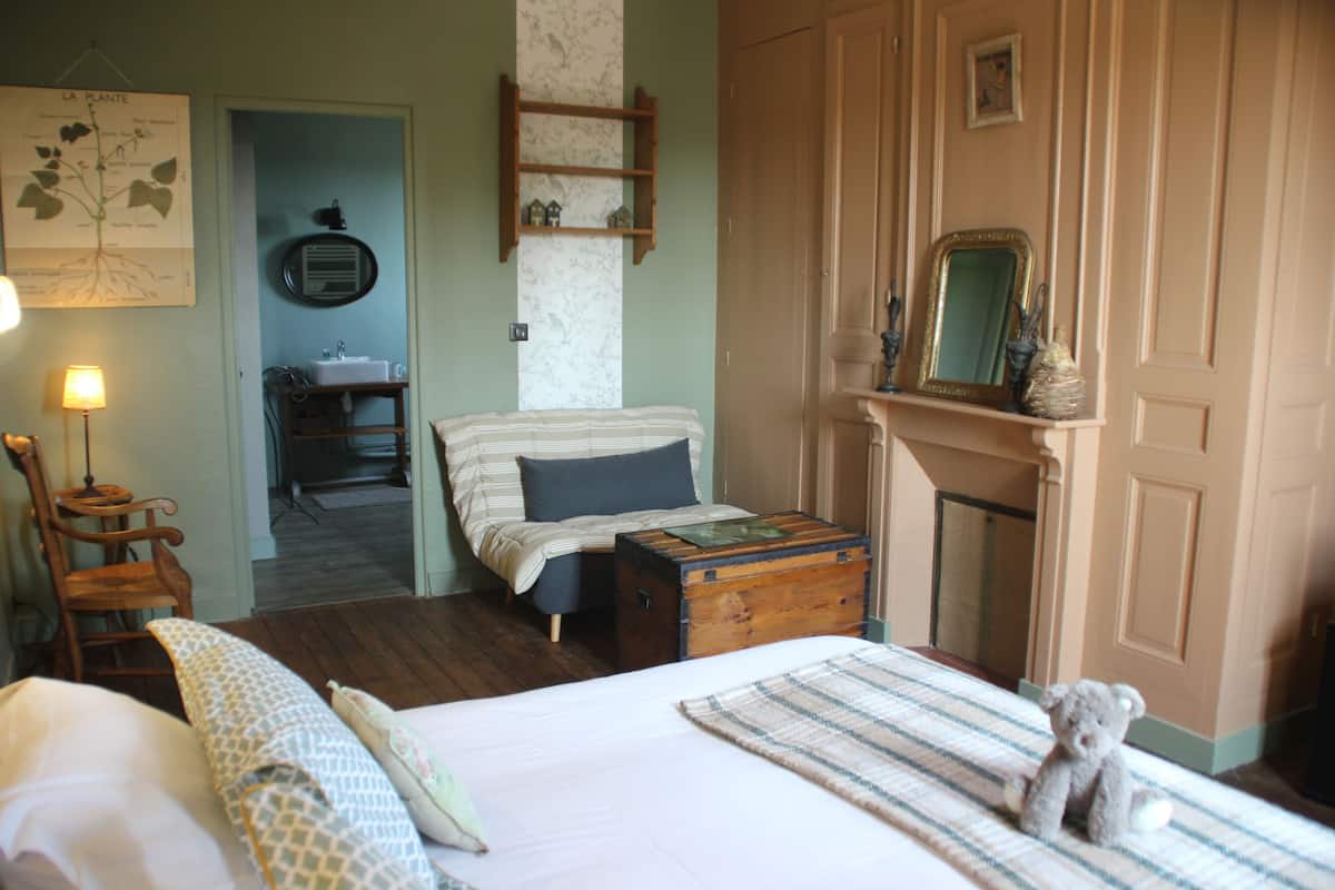 Quarto com cama, cómoda, espelho, lareira e um pequeno baú de madeira.