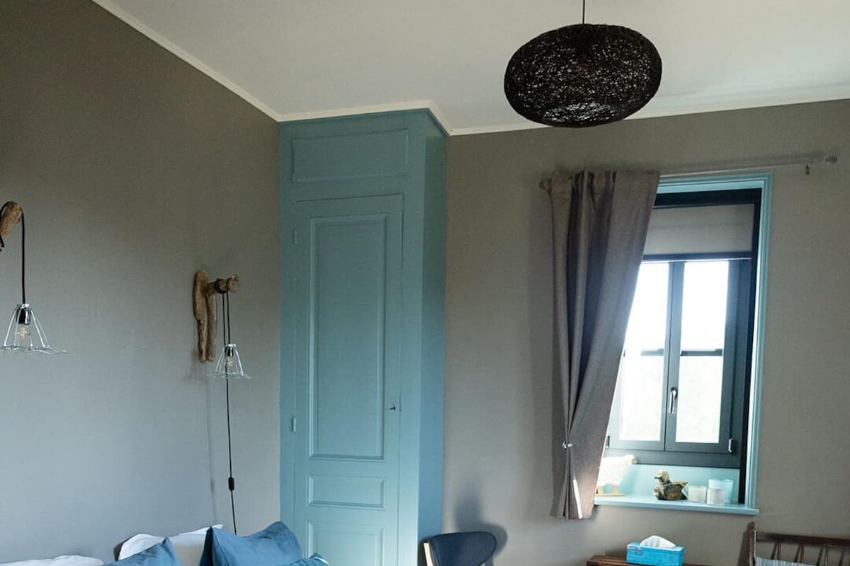 Quarto com cama grande, piso de madeira, janela com cortinas, uma cadeira e mesa de cabeceira.