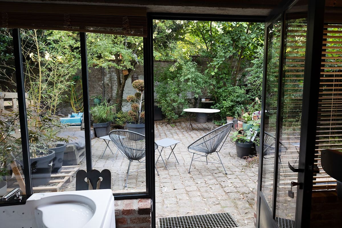 Casa de banho com uma grande janela que dá para um jardim com plantas em vasos e mobiliário de exterior.