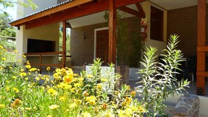 Um espaço ao ar livre coberto com estrutura de madeira, um banco de pedra e diversas plantas e flores.