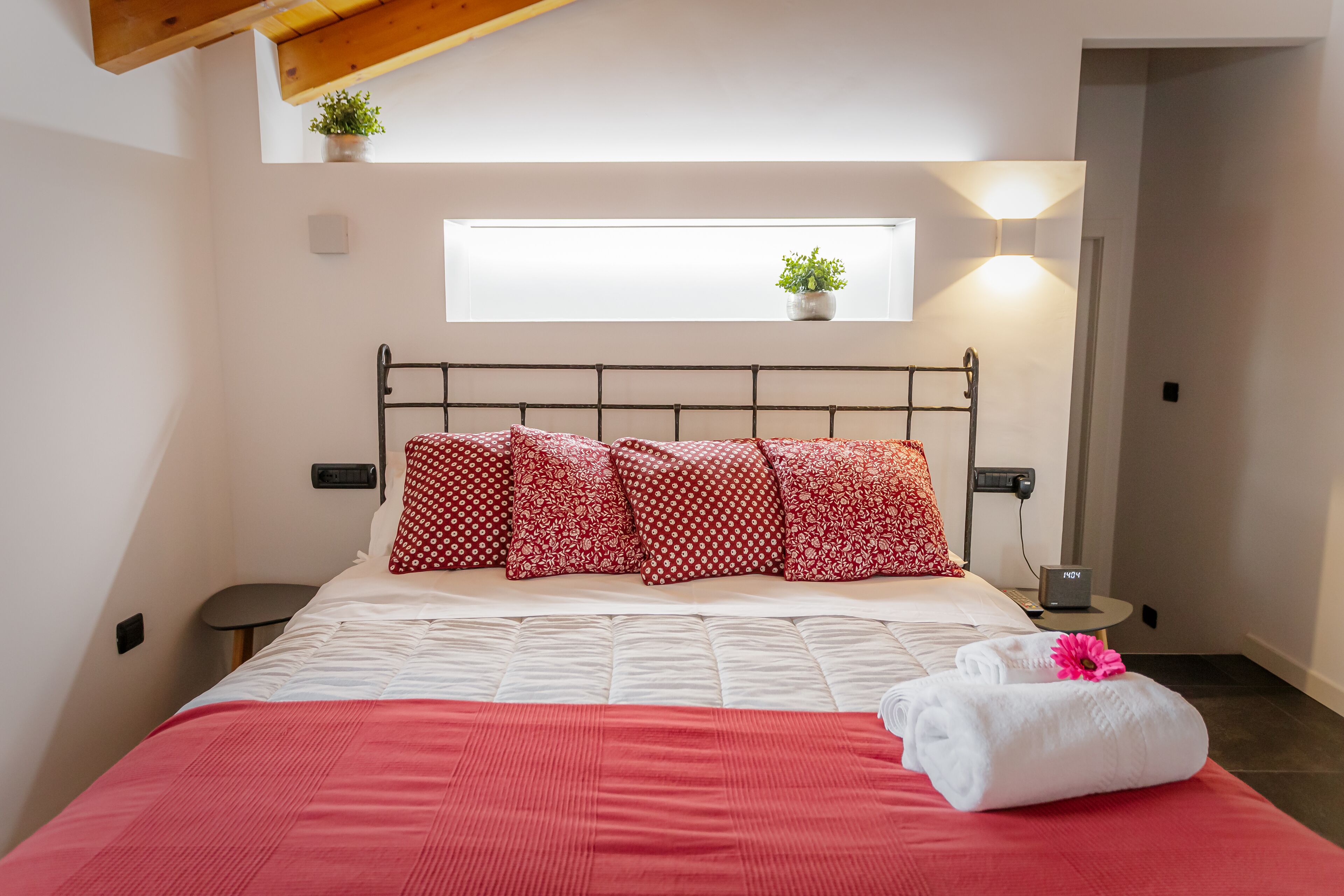 Cama bem arrumada com roupa de cama vermelha e branca, uma pequena mesa de cabeceira com abajur e uma janela com uma planta.