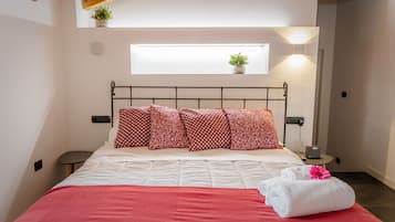Cama bem arrumada com roupa de cama vermelha e branca, uma pequena mesa de cabeceira com abajur e uma janela com uma planta.