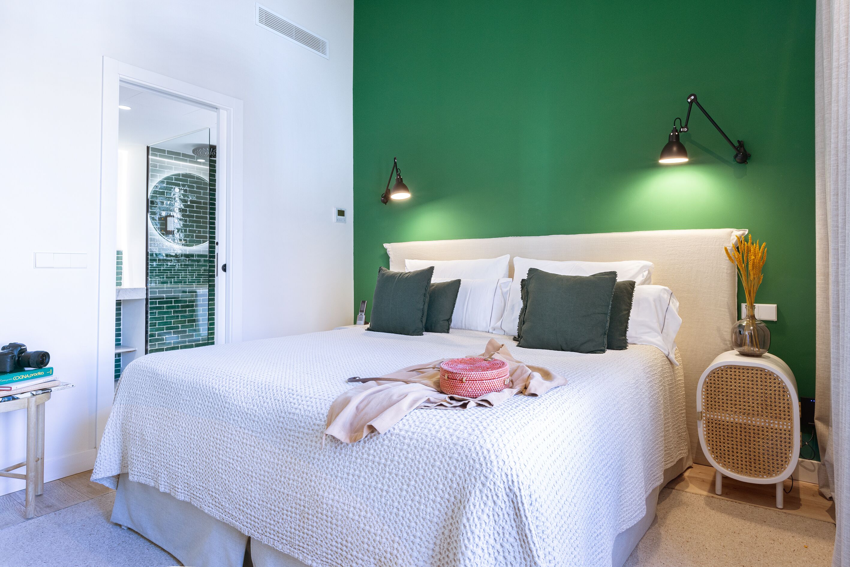 Cama bem arrumada com edredom branco, duas luminárias de cabeceira e uma pequena mesa de cabeceira de vime com um vaso.