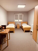 Quarto com duas camas de solteiro, uma escrivaninha, uma cadeira, radiador, janela e guarda-roupa.