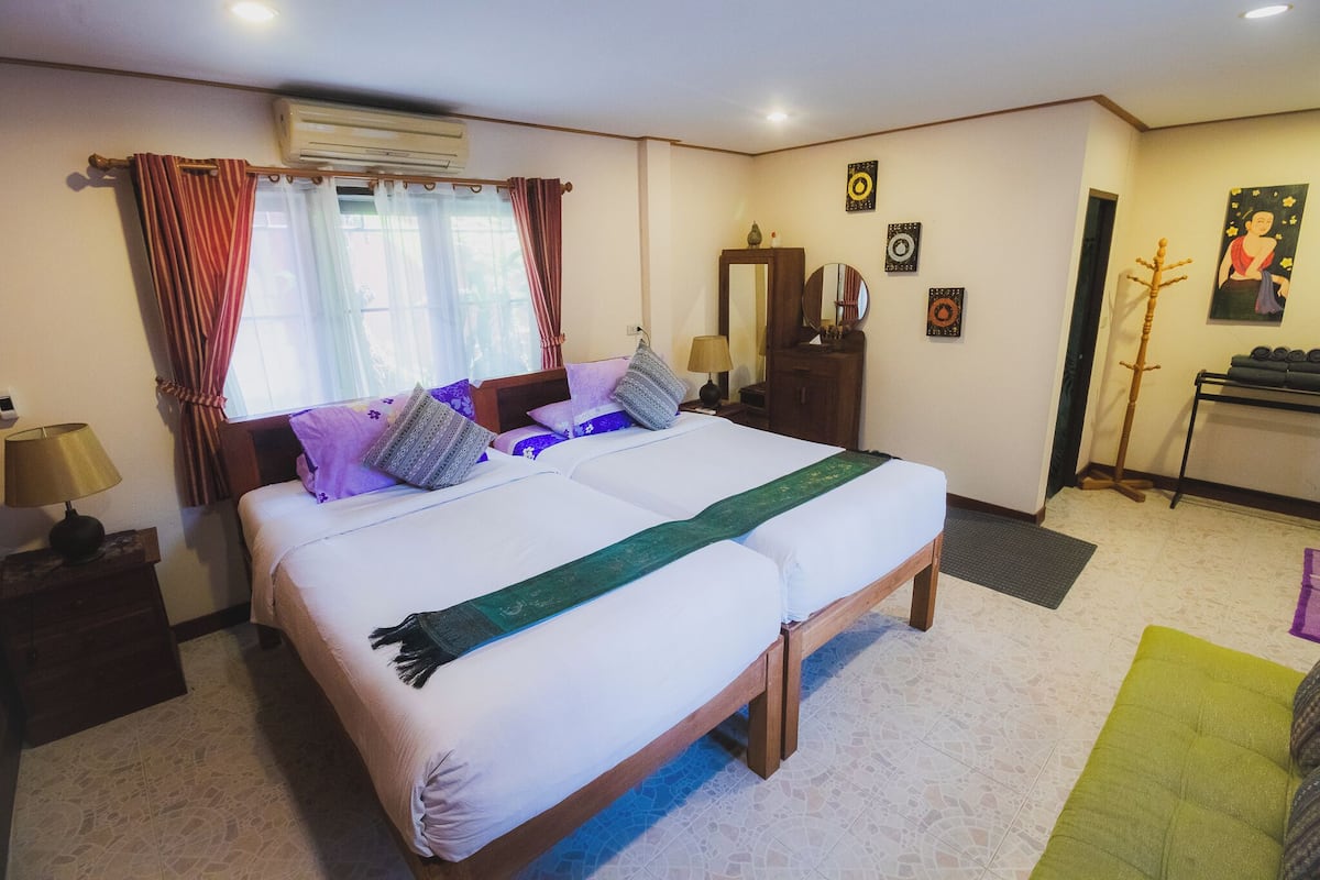 Uma cama de casal com um cobertor verde, uma mesa de cabeceira com abajur, um aparador de madeira, um espelho e um quadro na parede.