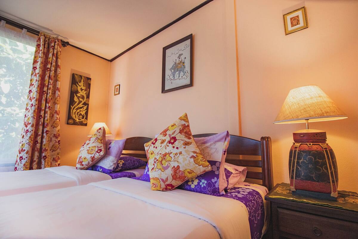 Cama bem arrumada com roupa de cama floral e roxa, mesa de cabeceira com abajur e quadro na parede.