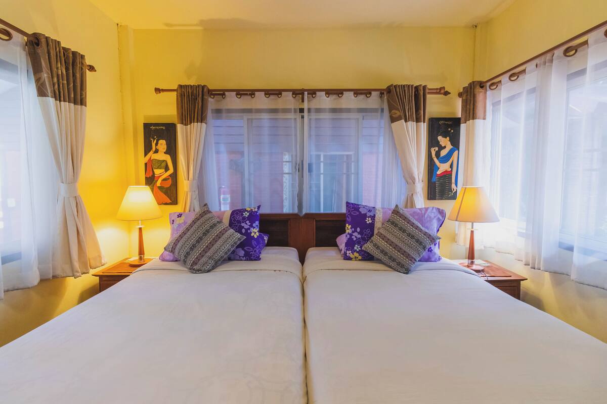 Quarto de hotel com duas camas, uma janela grande com cortinas e duas luminárias fixadas na parede.