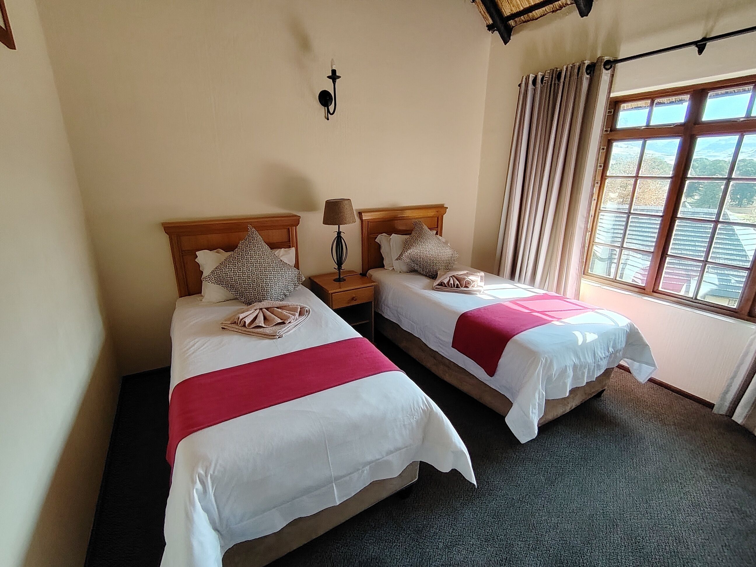 Quarto de hotel com duas camas, uma mesa de cabeceira com abajur, uma janela com cortinas e vista para o exterior.