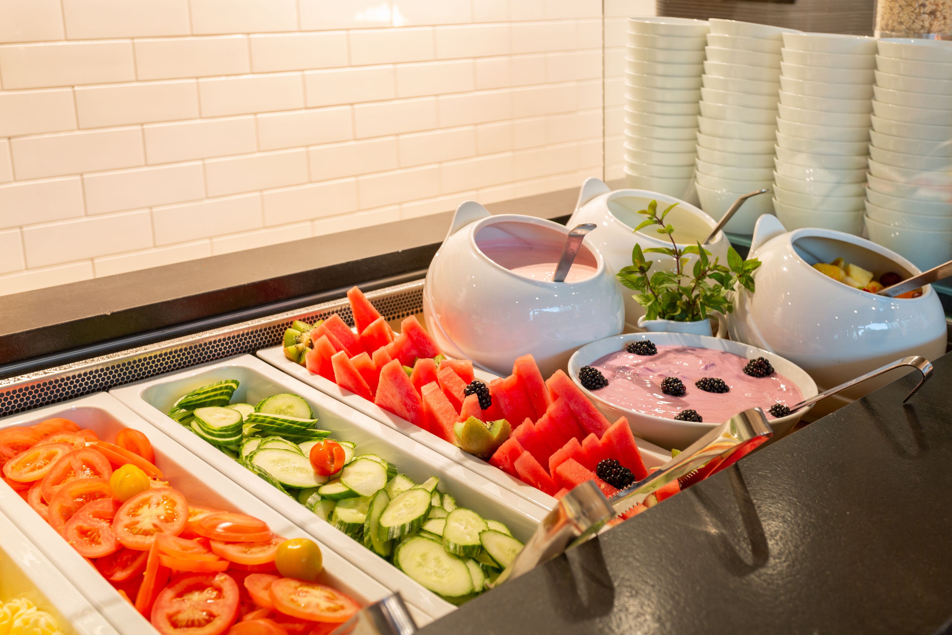 Buffet com diversas frutas e legumes, incluindo tomates, pepinos e melancia, além de potes de iogurte e outros itens.