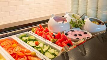 Buffet com diversas frutas e legumes, incluindo tomates, pepinos e melancia, além de potes de iogurte e outros itens.
