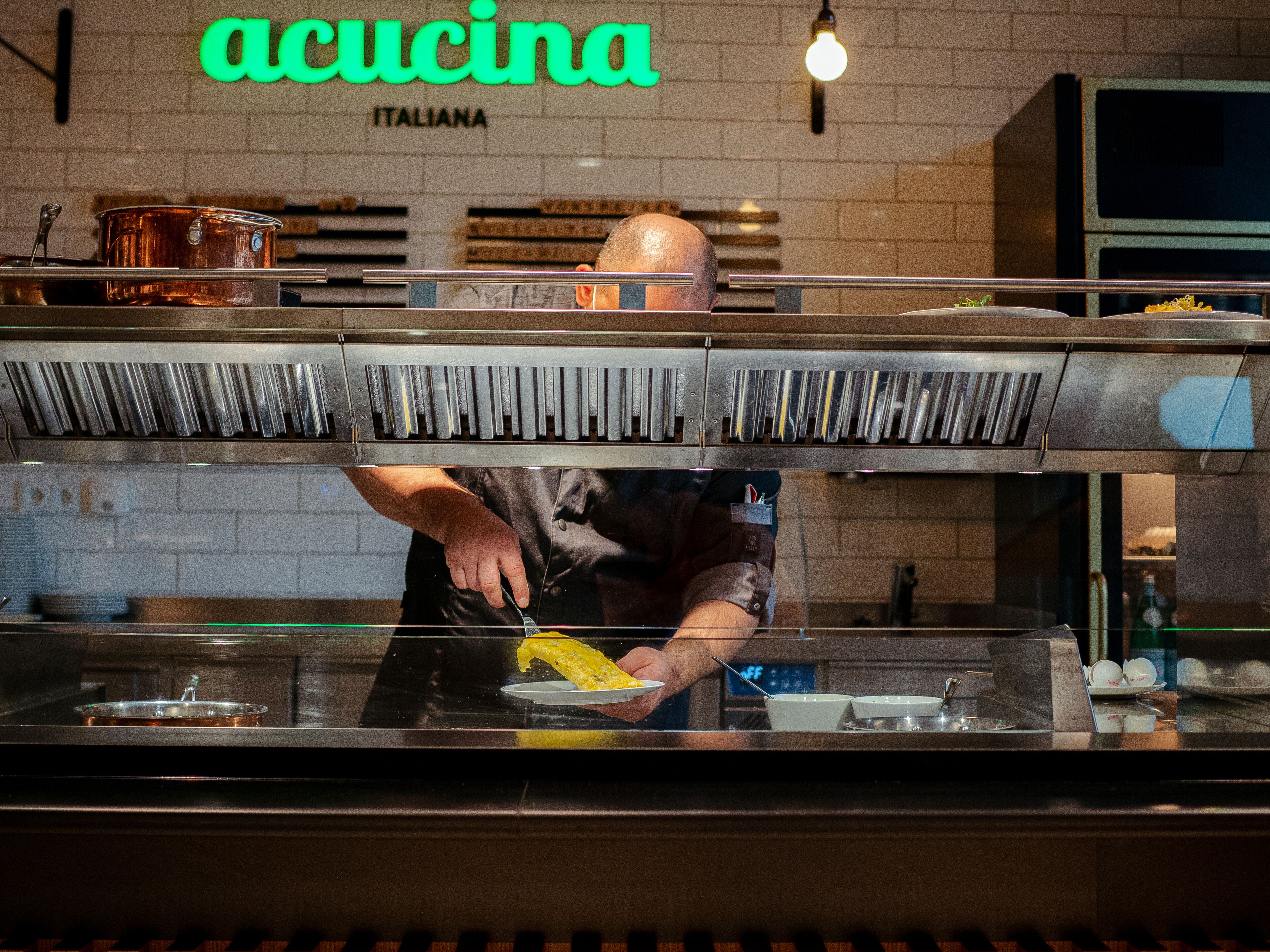 Uma bancada de cozinha com um chef preparando comida, um letreiro com a inscrição "acucina ITALIANA" e utensílios de cozinha.
