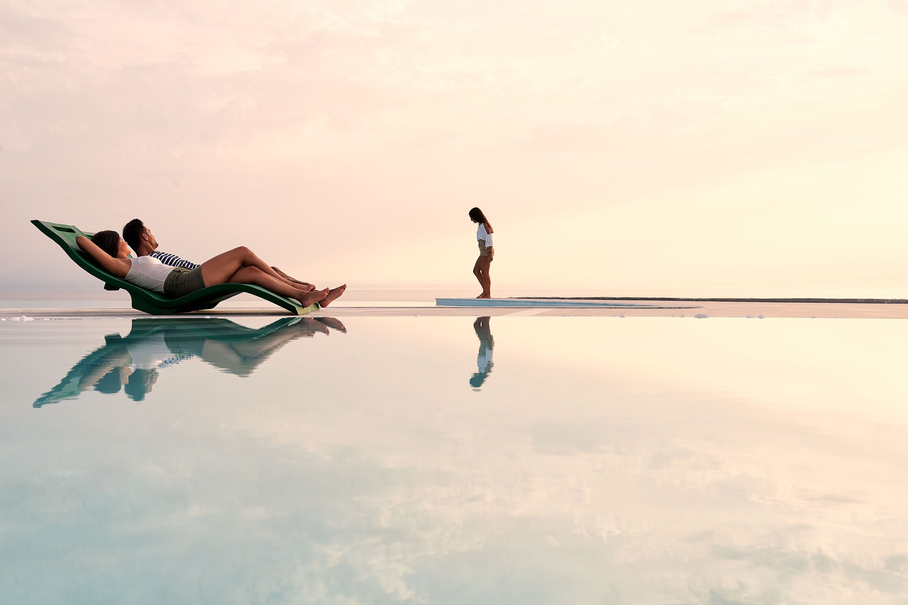 Um casal relaxando em uma espreguiçadeira perto de uma piscina espelhada, com vista para o oceano.