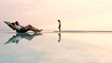 Um casal relaxando em uma espreguiçadeira perto de uma piscina espelhada, com vista para o oceano.