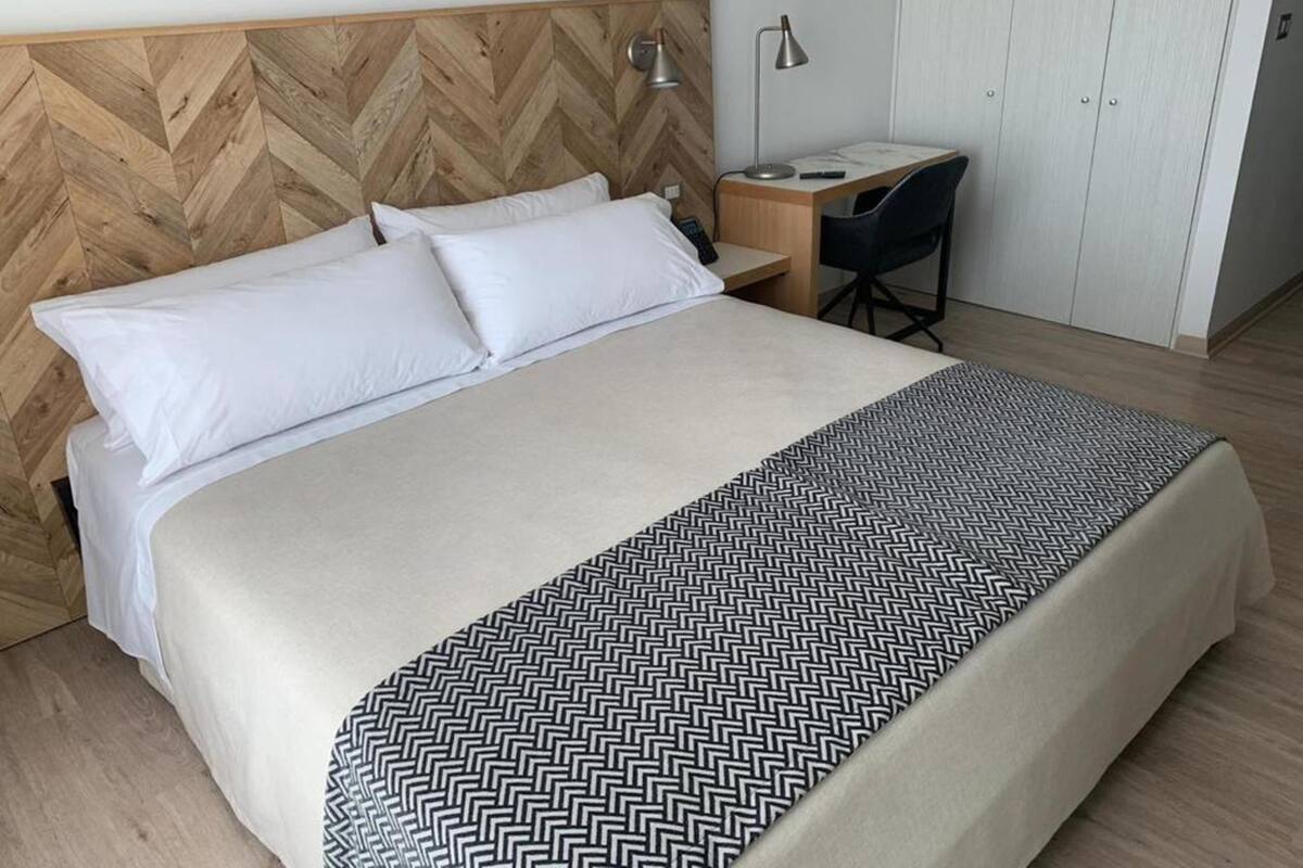 Uma cama bem arrumada com roupa de cama branca, cabeceira de madeira, escrivaninha com cadeira e um guarda-roupa.