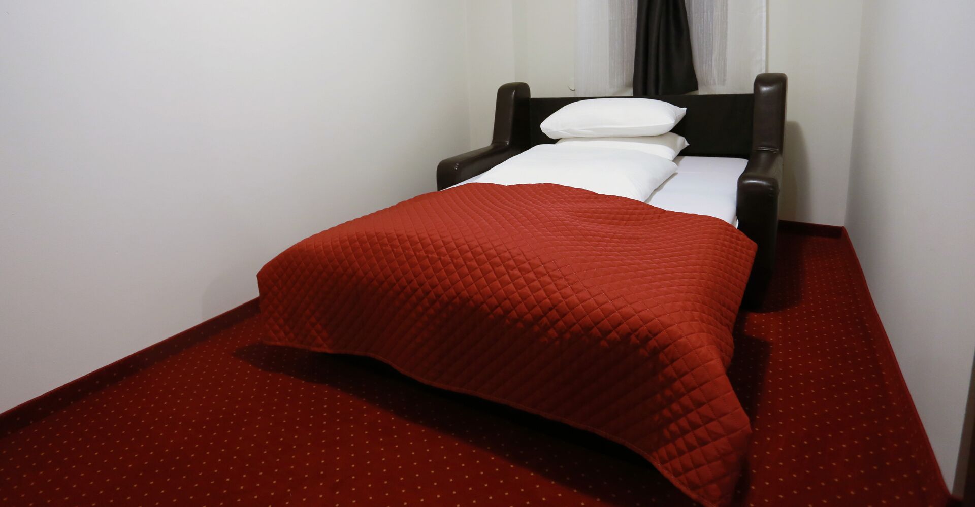 Quarto pequeno com cama de solteiro, edredom acolchoado vermelho e travesseiros brancos.