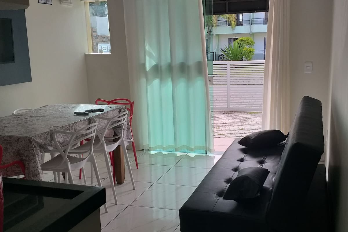 Quarto pequeno e bem iluminado, com mesa e cadeiras de jantar, um sofá e uma porta de vidro deslizante para área externa.