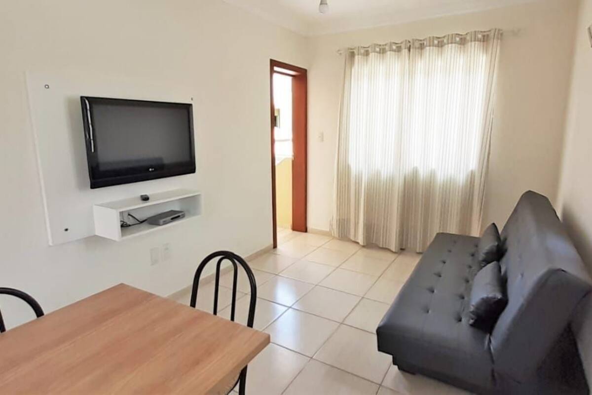 Uma sala de estar com televisão na parede, um sofá, uma mesa de jantar com cadeiras e uma janela com cortinas.