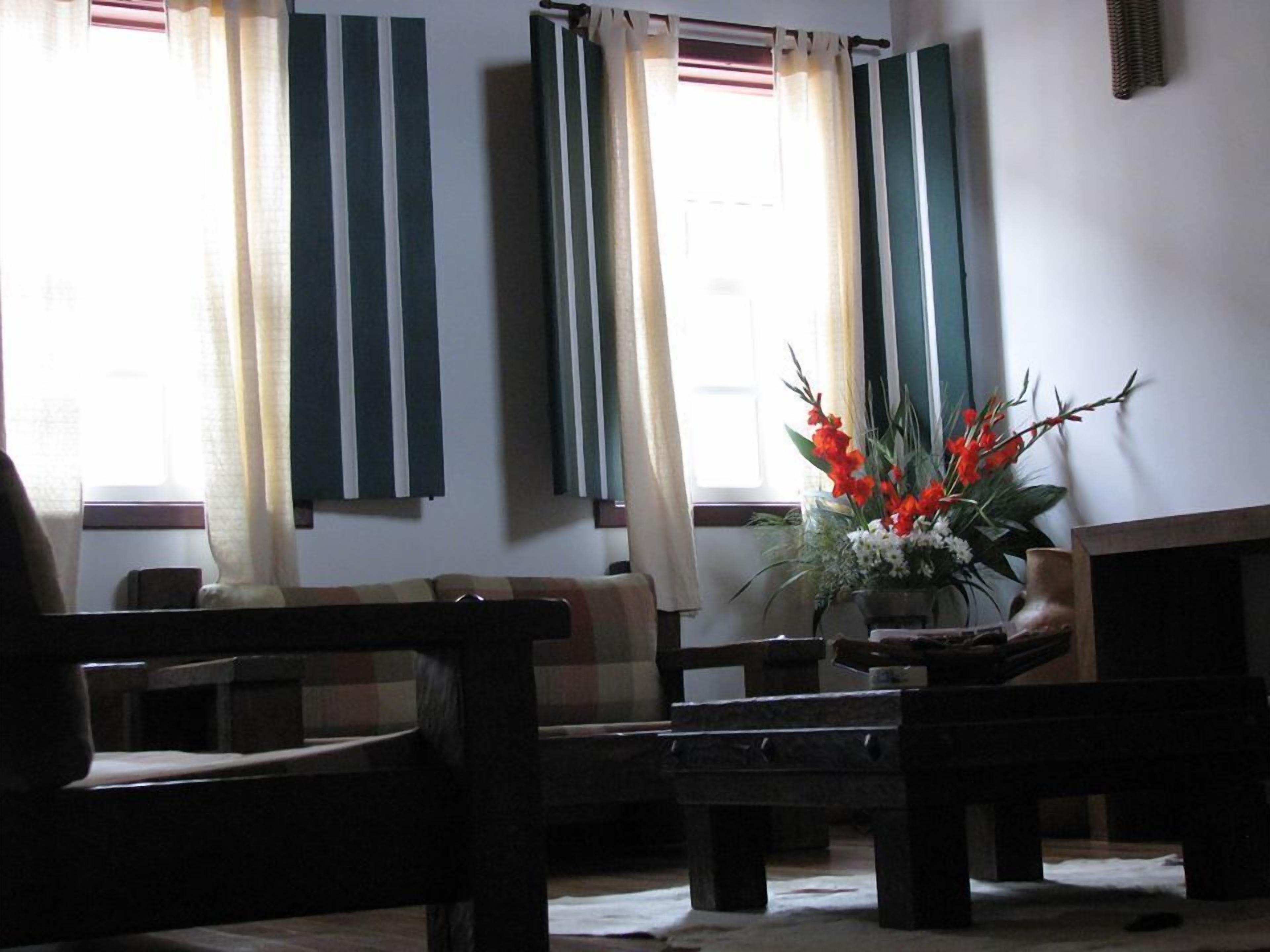 Sala de estar com sofá, duas poltronas, uma mesa de centro e um vaso com flores. Há cortinas nas janelas e uma planta no parapeito.
