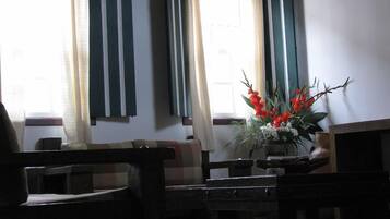 Sala de estar com sofá, duas poltronas, uma mesa de centro e um vaso com flores. Há cortinas nas janelas e uma planta no parapeito.