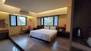 Quarto de hotel moderno com uma cama grande, duas mesas de cabeceira, uma escrivaninha e uma cadeira. Possui duas janelas com cortinas, um aparelho de ar condicionado e uma máquina de café.