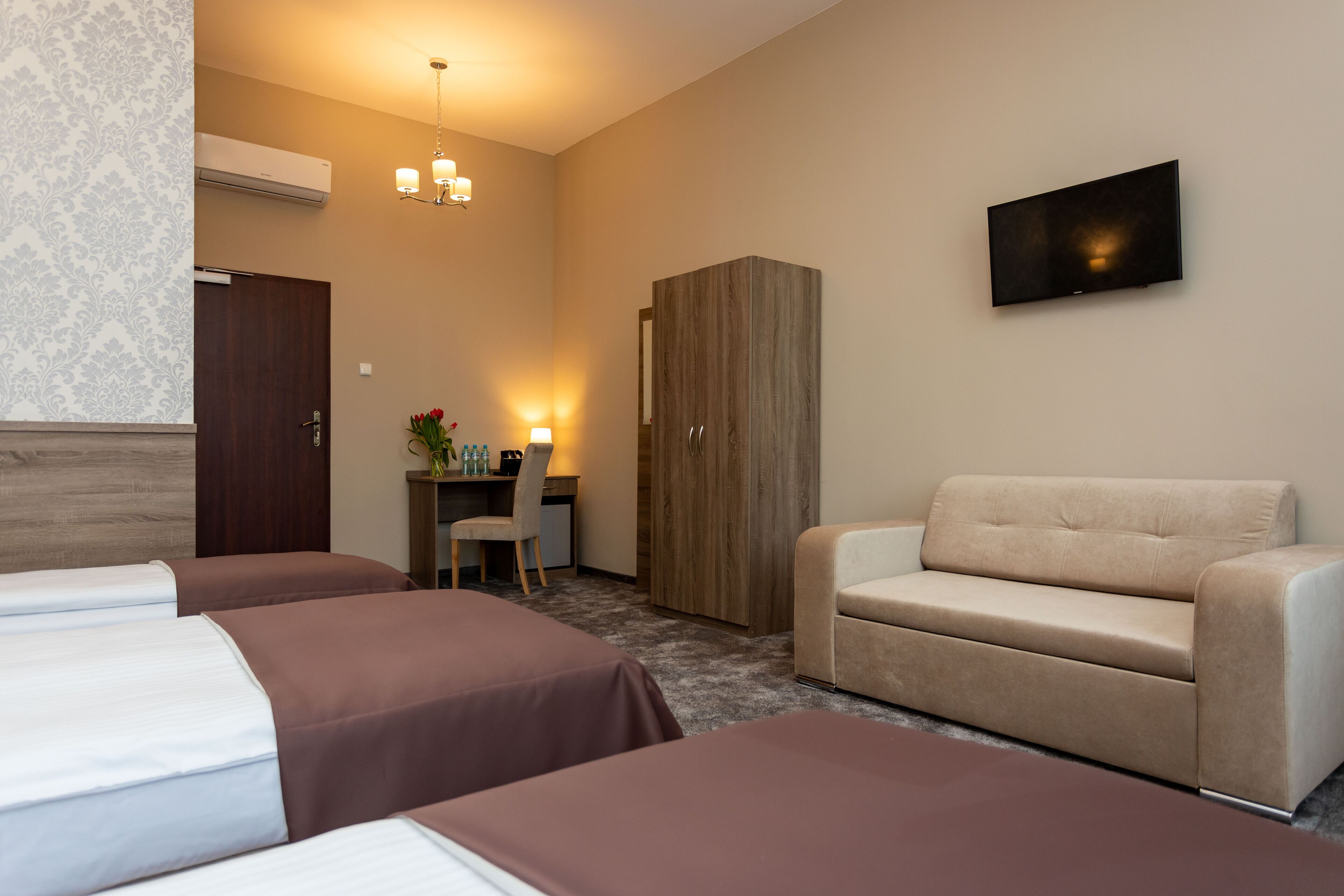 Quarto de hotel com duas camas, um sofá, uma mesa de trabalho com cadeira, televisão e guarda-roupa.