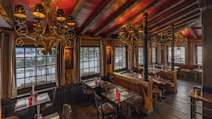 Restaurante com vigas de madeira, lustres e janelas amplas. As mesas estão dispostas com guardanapos vermelhos e copos.