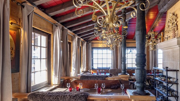Um quarto espaçoso com teto alto, vigas de madeira e um grande candelabro. Há mesas com copos vermelhos e um banco forrado com pele.
