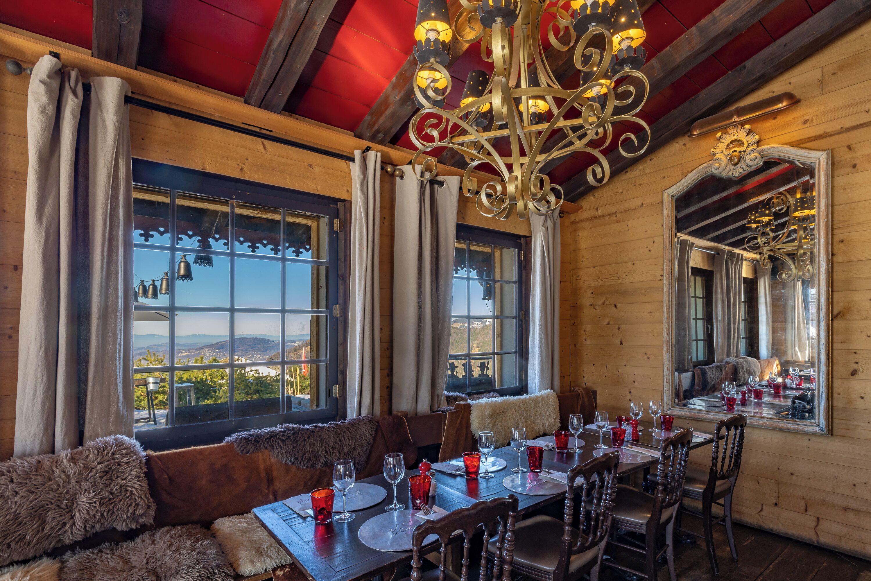 Interior em madeira com uma mesa de jantar posta para quatro, janelas amplas com cortinas e um candelabro.