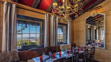 Interior em madeira com uma mesa de jantar posta para quatro, janelas amplas com cortinas e um candelabro.