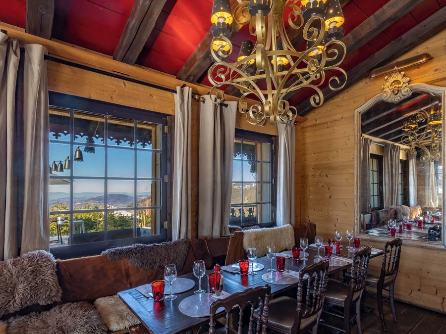 Interior em madeira com uma mesa de jantar posta para quatro, janelas amplas com cortinas e um candelabro.