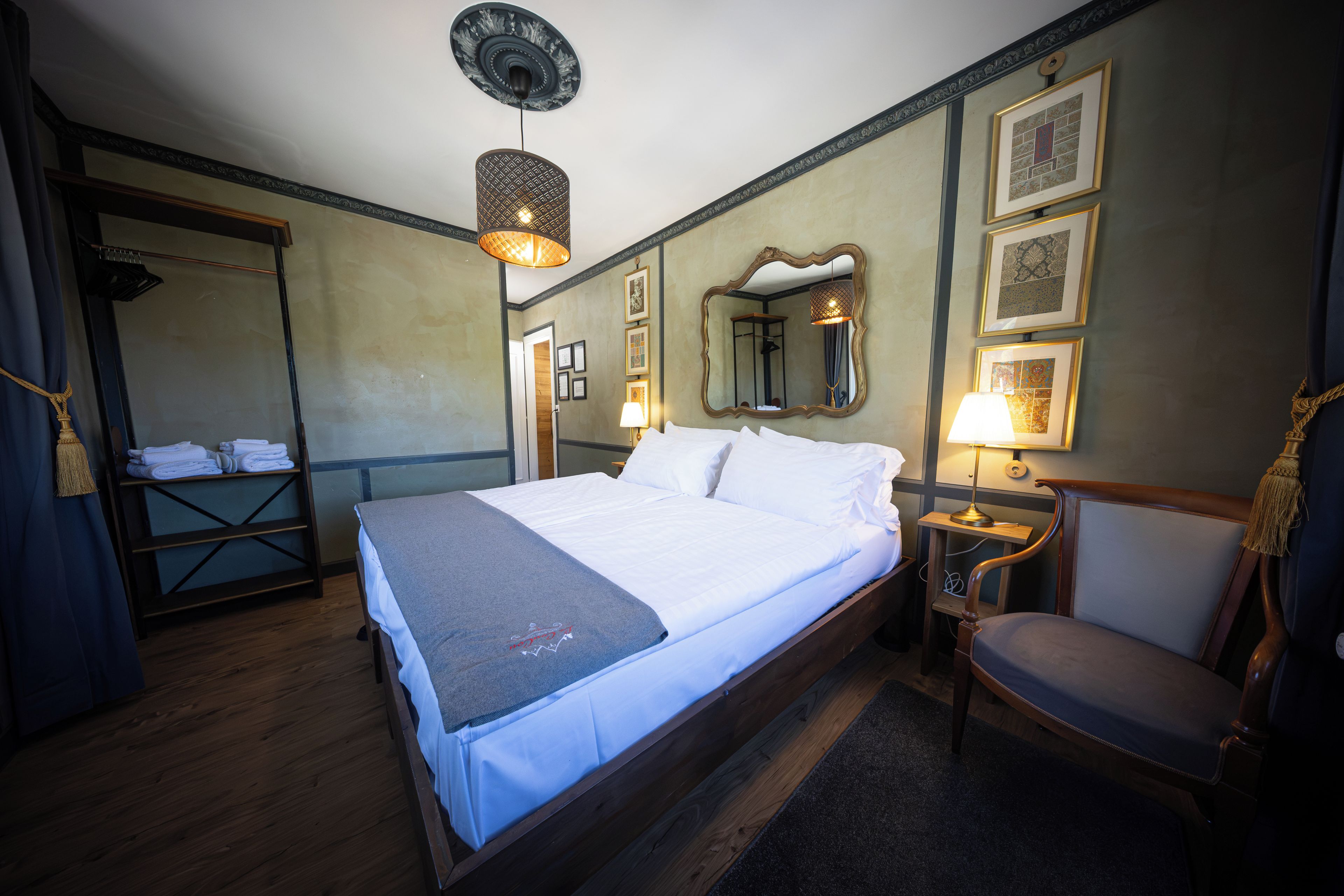 Quarto de hotel com uma cama grande, uma cadeira, uma escrivaninha com abajur, espelho e guarda-roupa.