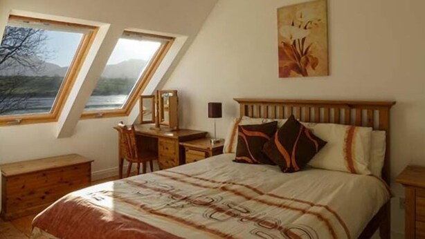 Quarto com cama, escrivaninha e duas janelas com vista para a paisagem.