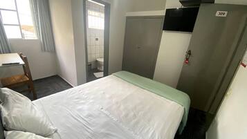 Quarto de hotel com cama, escrivaninha, cadeira, guarda-roupa e televisão.