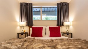Quarto com cama grande, duas luminárias de cabeceira, janela com cortinas e vista para área verde lá fora.