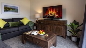 Uma sala de estar com um sofá escuro, uma mesa de centro com uma fruteira, uma televisão exibindo uma lareira e um vaso de plantas.
