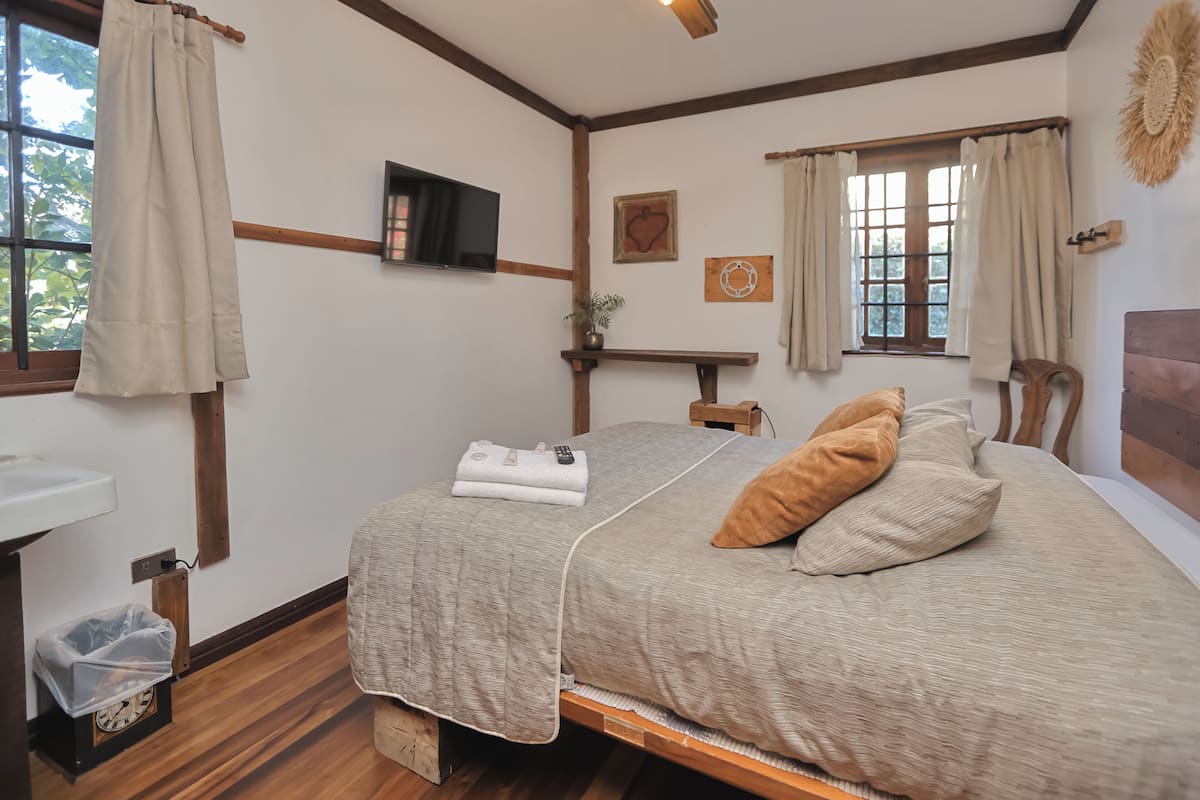 Quarto com cama, televisão, janela com cortinas, piso de madeira e pequena mesa de cabeceira.