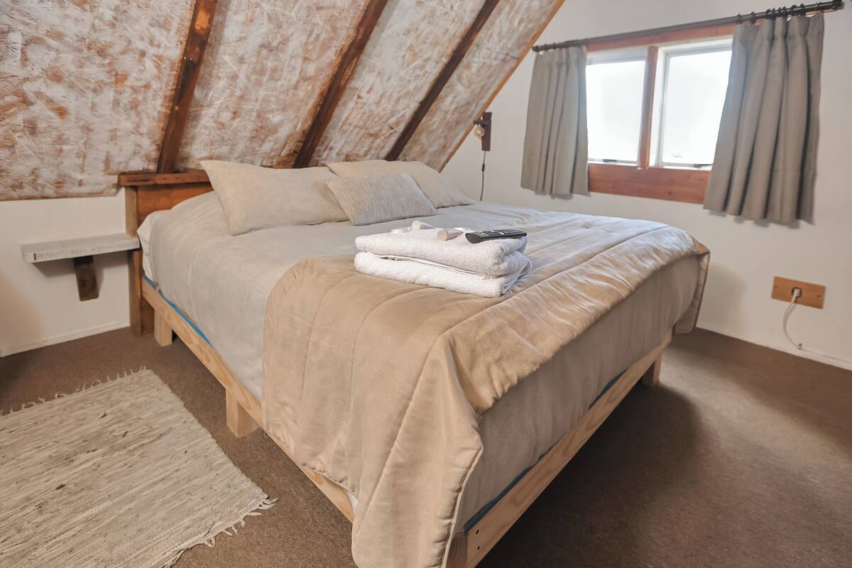 Um quarto com teto inclinado, uma cama com manta bege, uma mesinha de cabeceira, uma janela com cortinas e uma tomada na parede.