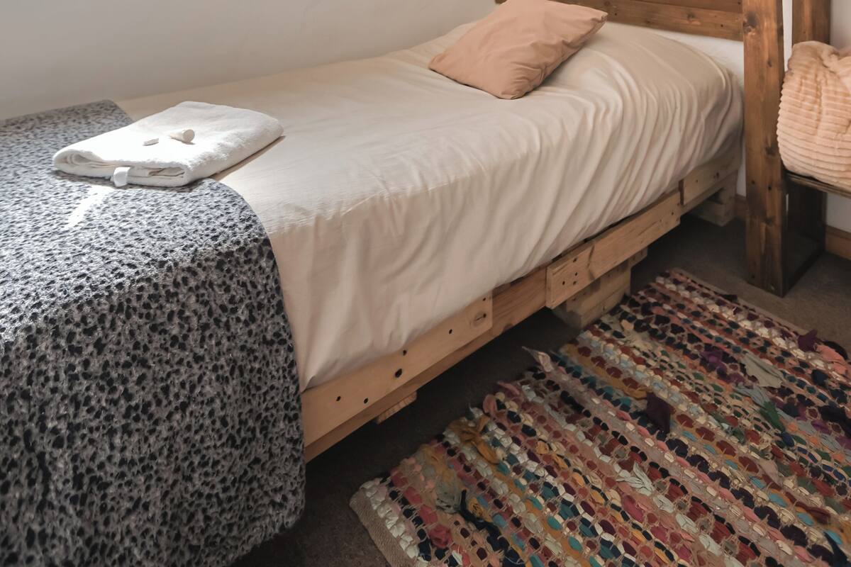 Quarto com cama, janela, teto de madeira e tapete colorido.