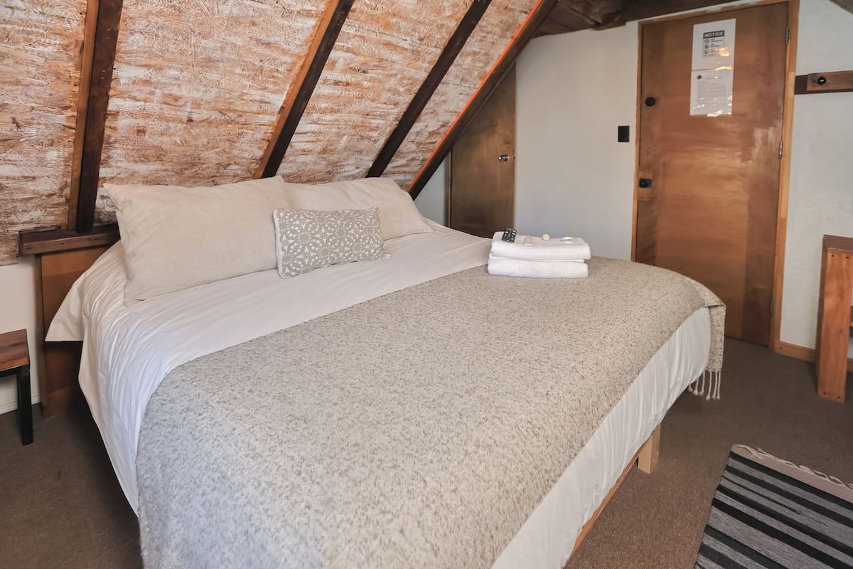 Quarto com teto inclinado, cama com roupa de cama branca, porta de madeira e um pequeno banco.