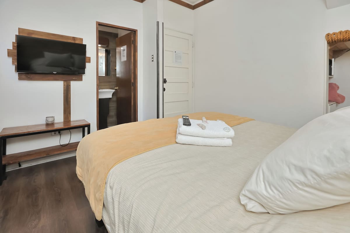 Quarto com uma cama, TV fixada na parede, um banco de madeira e porta que dá para um banheiro.
