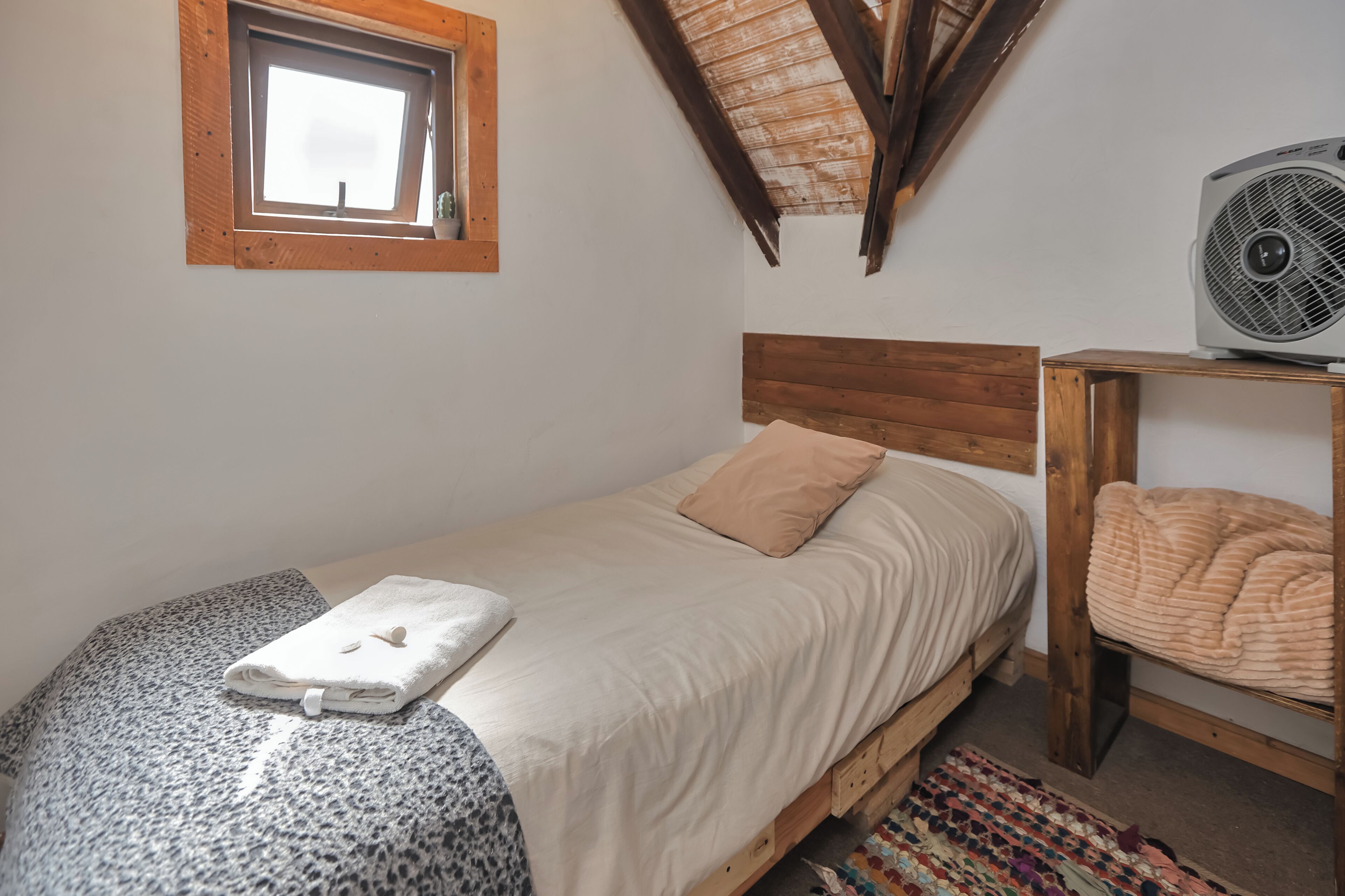 Uma cama de solteiro com cabeceira de madeira, uma janela pequena, um ventilador e um banquinho de madeira com um cobertor.