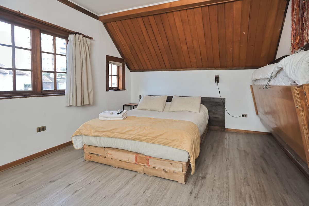 Quarto com teto de madeira, cama com estrutura de madeira, janela com cortinas e uma pequena mesa de cabeceira.