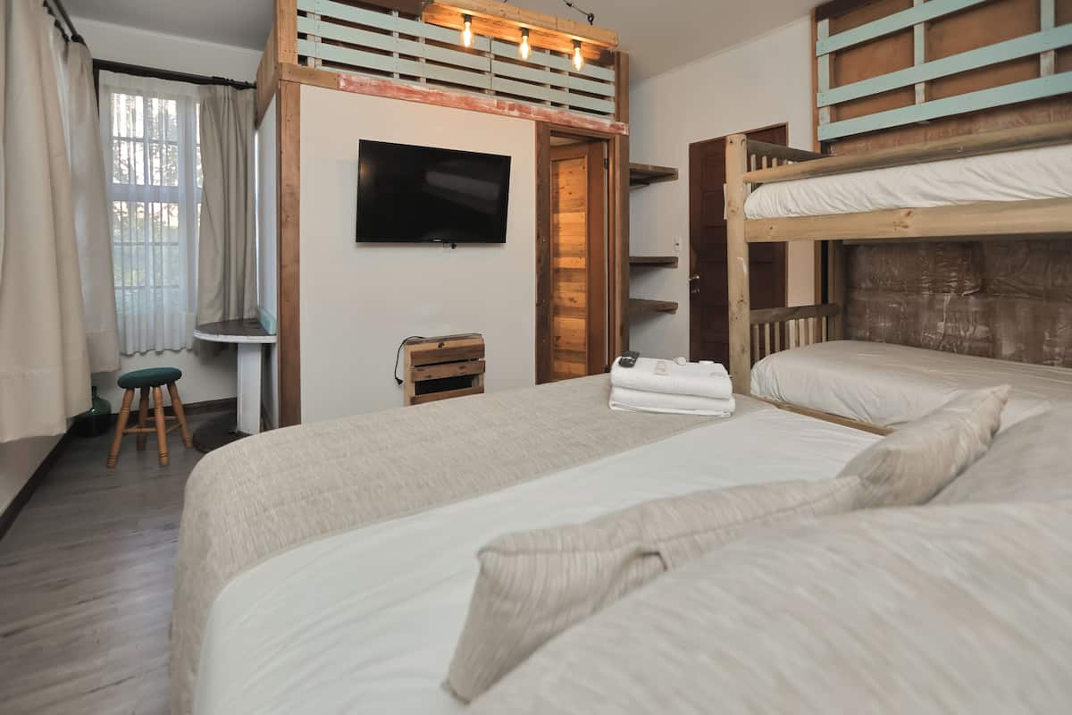 Quarto com beliches, televisão, prateleira de madeira e janela com cortinas.
