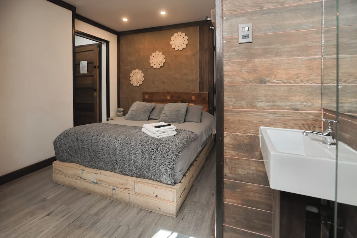 Quarto com estrutura de cama de madeira, manta cinza e toalha branca. Há uma parede de madeira com desenhos circulares decorativos e uma pia branca com espelho.